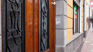 Uma porta de madeira com detalhes metálicos elaborados, uma rua de calçada de tijolos e um edifício com a placa 'Hotel'.