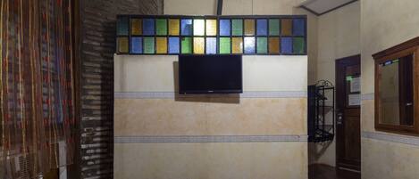 Quarto com televisão fixada na parede sobre um padrão de azulejos, uma porta de madeira e uma estante de metal com objetos no lado direito.