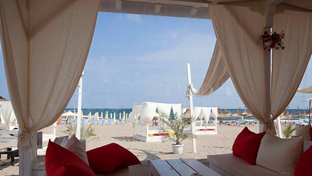 Um espaço de relaxamento à beira-mar com mobiliário branco e almofadas vermelhas, com vista para uma praia arenosa com guarda-sóis e cadeiras de praia.