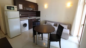 Uma cozinha compacta com micro-ondas, frigorífico e máquina de lavar roupa. Possui uma área de refeições com mesa e cadeiras, e um sofá com almofadas.