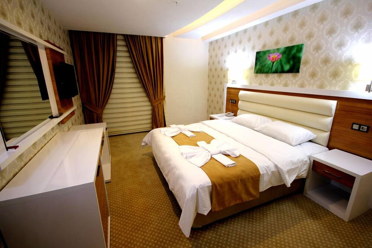 Quarto de hotel com cama, escrivaninha, cadeira, mesa de cabeceira, televisão e janela com cortinas.