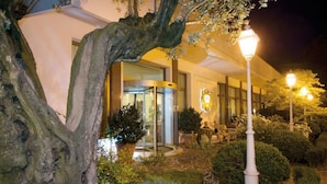 Entrada de hotel com porta giratória, caminho bem iluminado e área de jardim com poste de luz.