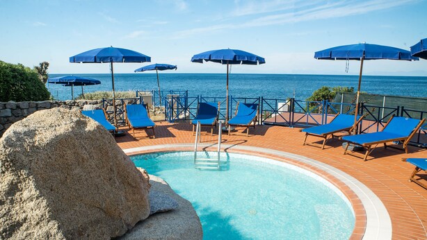 Área da piscina com cadeirões azuis e guarda-sóis, vista desobstruída do mar e uma rocha ornamental.