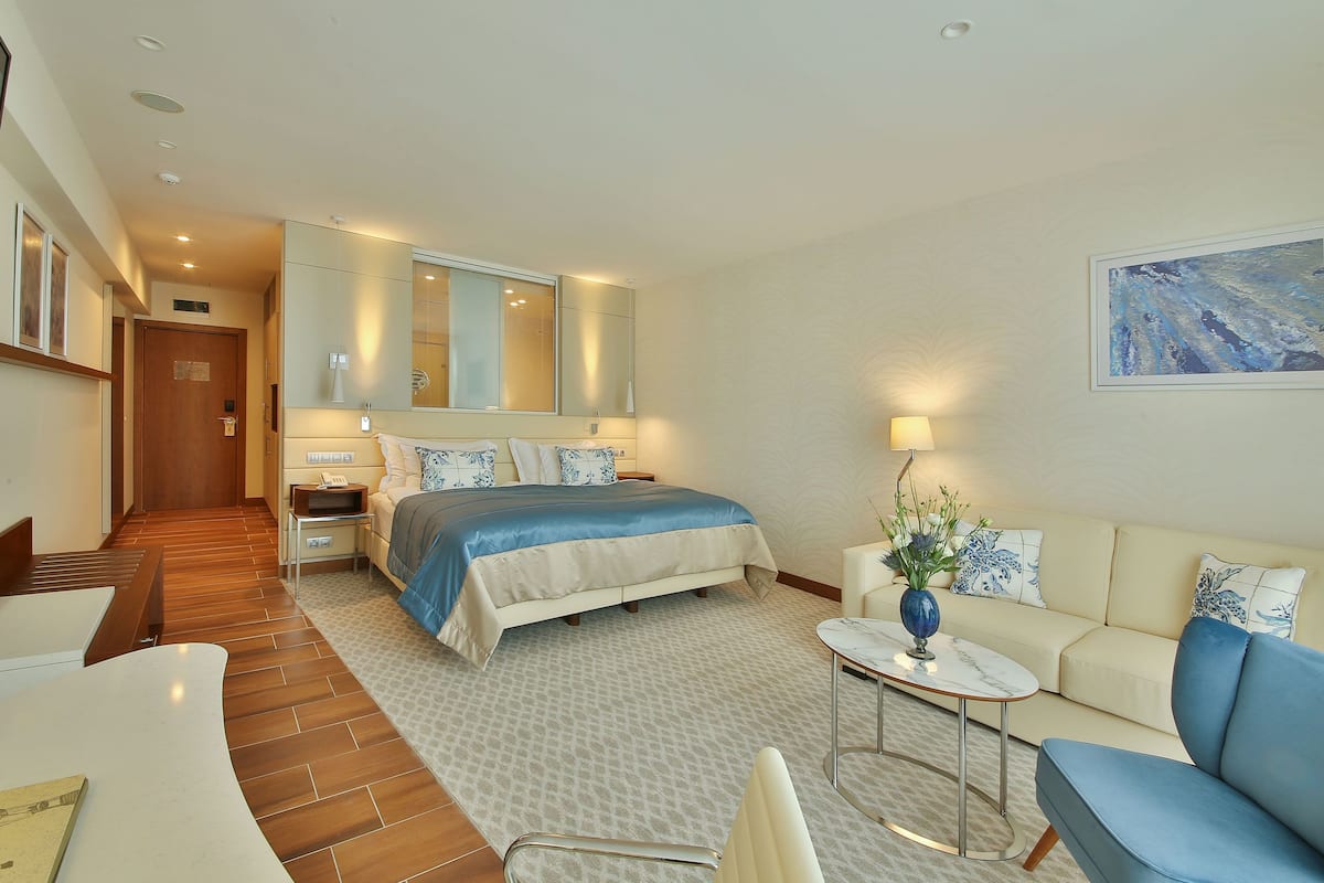 Quarto de hotel com cama, sofá, mesinha e cadeira. Piso de madeira e tapete estampado.