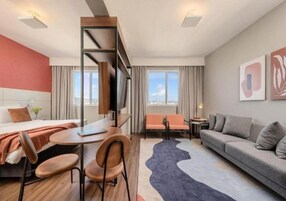 Quarto de hotel com cama, escrivaninha, sofá e cadeira. Há uma janela com cortinas e vista para a cidade.