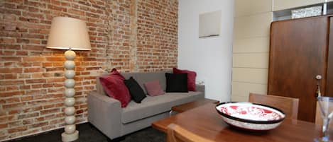 Uma sala de estar com parede de tijolos, um sofá cinza com almofadas vermelhas e pretas, uma mesa de jantar de madeira com uma tigela estampada em branco e vermelho, e um abajur branco.