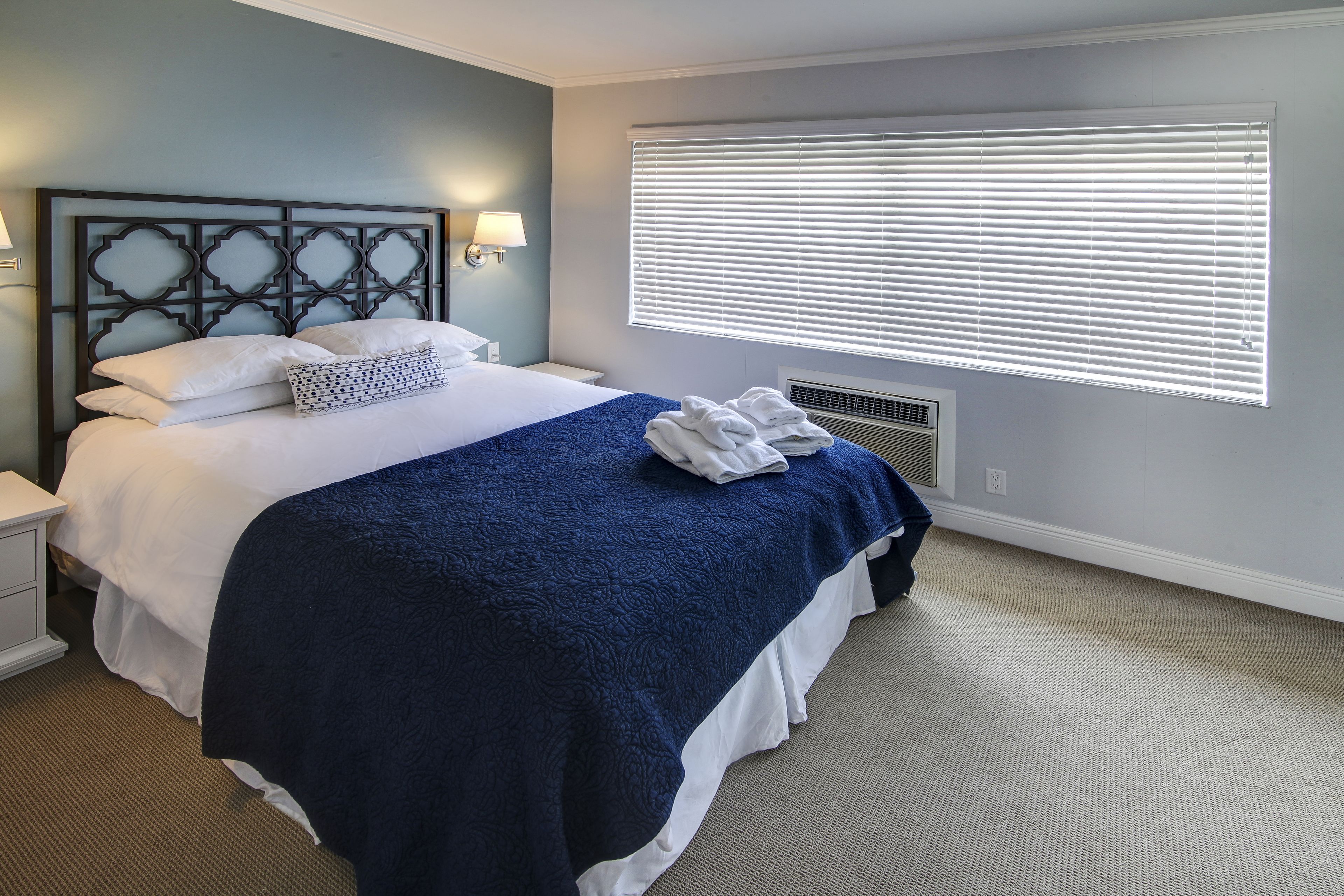 Uma cama bem arrumada com colcha azul e branca, uma cabeceira decorativa e um abajur fixado na parede.