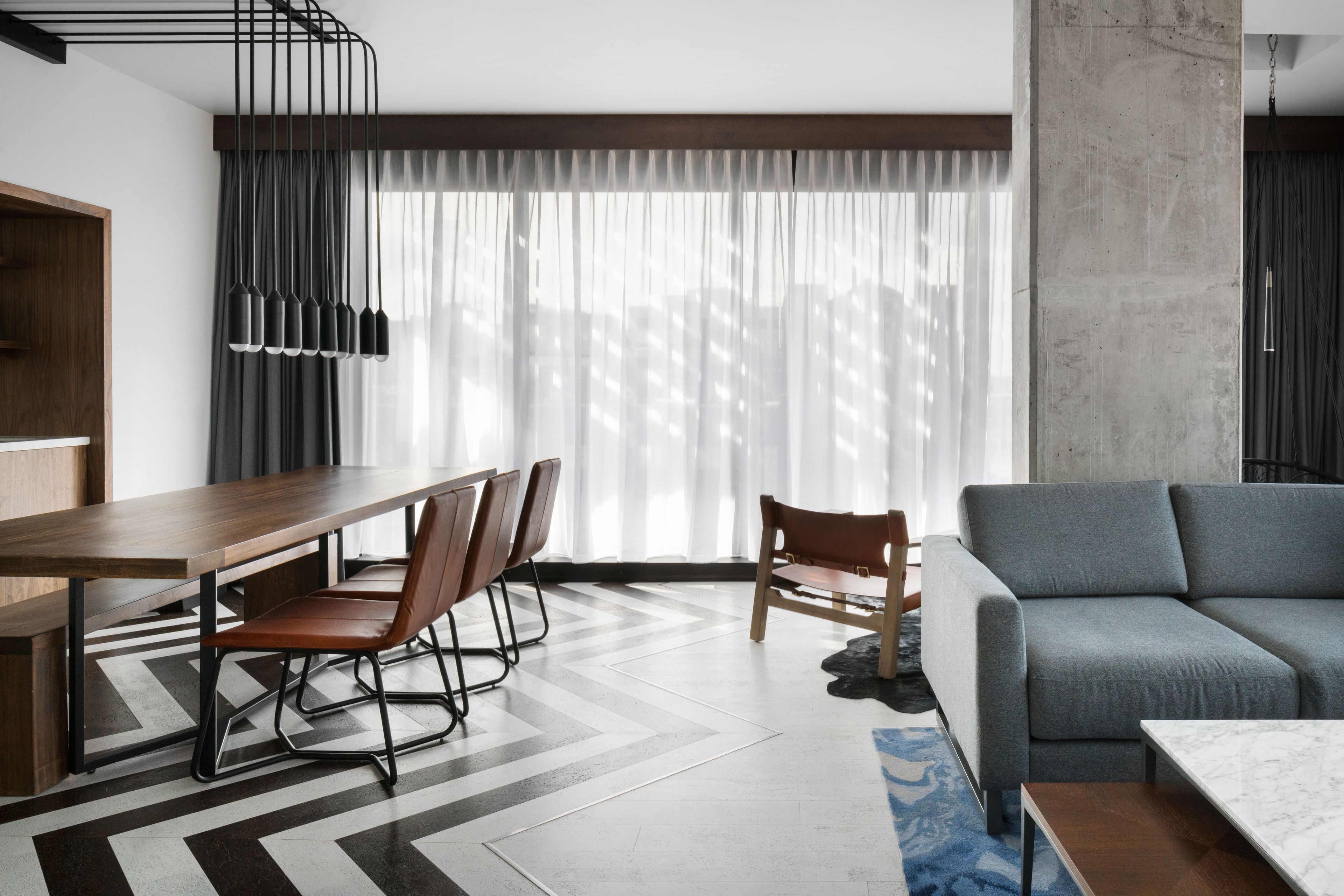 Uma sala de estar moderna com uma mesa de jantar de madeira, cadeiras, um sofá cinza e uma grande janela com cortinas leves.