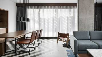 Uma sala de estar moderna com uma mesa de jantar de madeira, cadeiras, um sofá cinza e uma grande janela com cortinas leves.