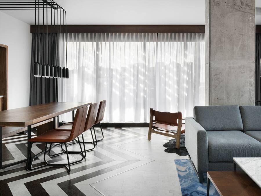 Uma sala de estar moderna com uma mesa de jantar de madeira, cadeiras, um sofá cinza e uma grande janela com cortinas leves.
