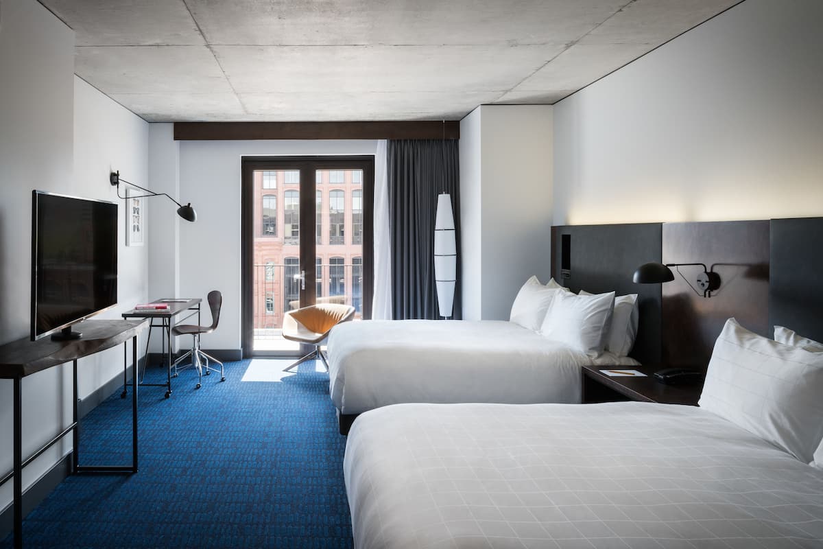 Quarto de hotel com duas camas, uma mesa de trabalho com televisão, uma cadeira e uma janela grande com vista para um edifício.