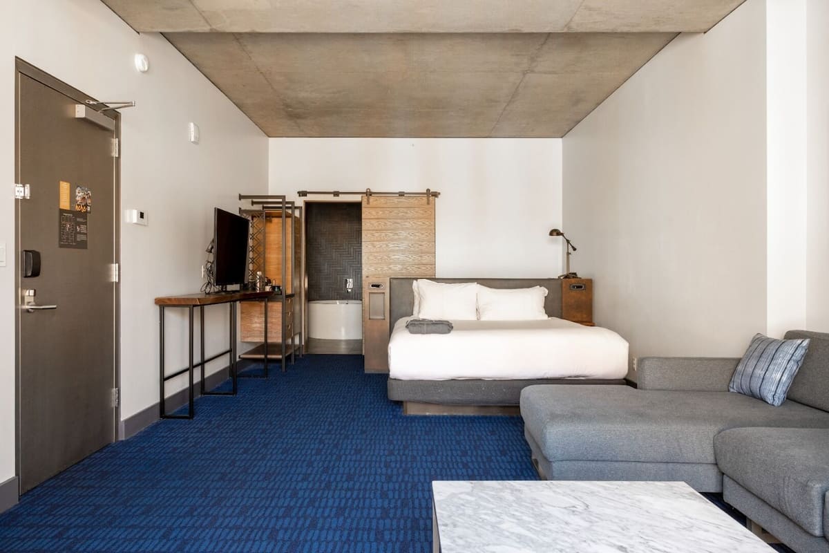 Quarto de hotel com cama, sofá, escrivaninha e computador, além de banheiro com banheira.