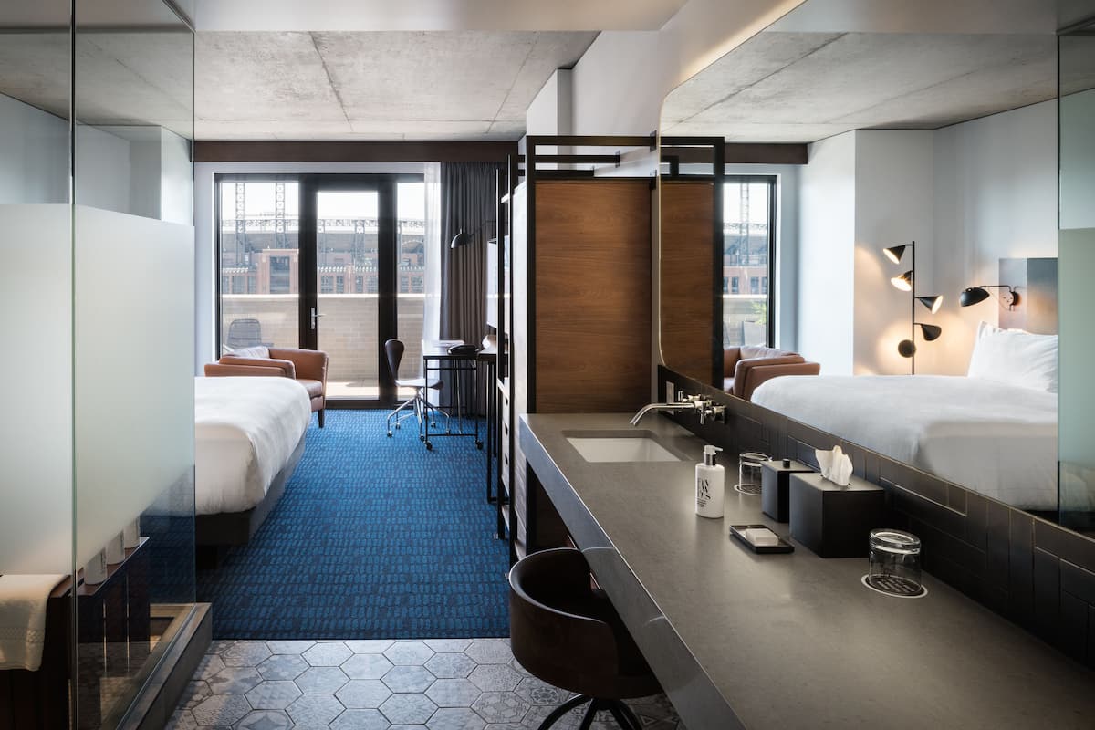 Quarto de hotel moderno com uma cama grande, uma escrivaninha, uma cadeira, banheiro com espelho e vista para a paisagem urbana.