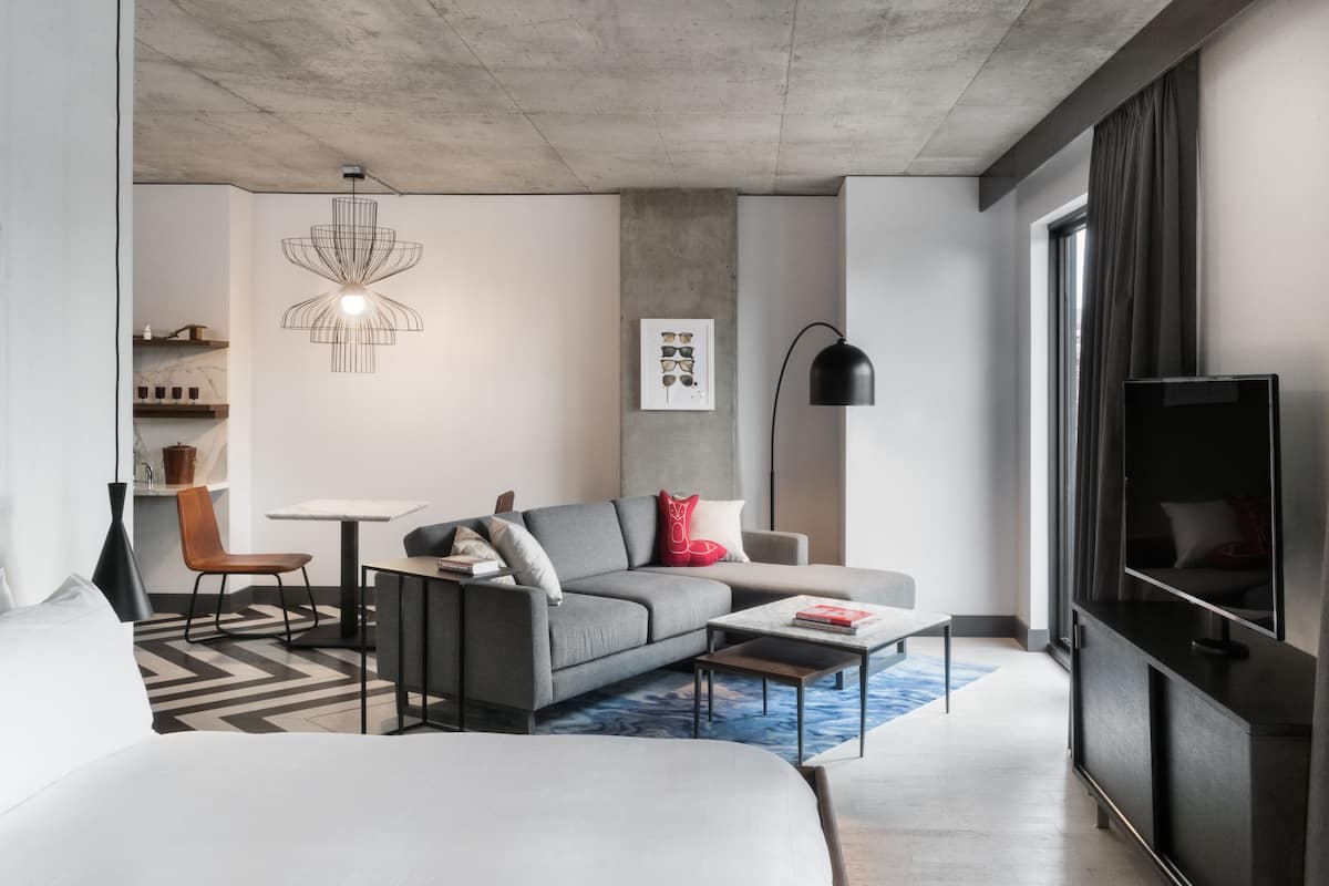 Uma sala de estar moderna com sofá, mesa de centro e televisão. Há uma área de jantar com mesa e cadeiras. O quarto possui teto de concreto e uma janela grande.