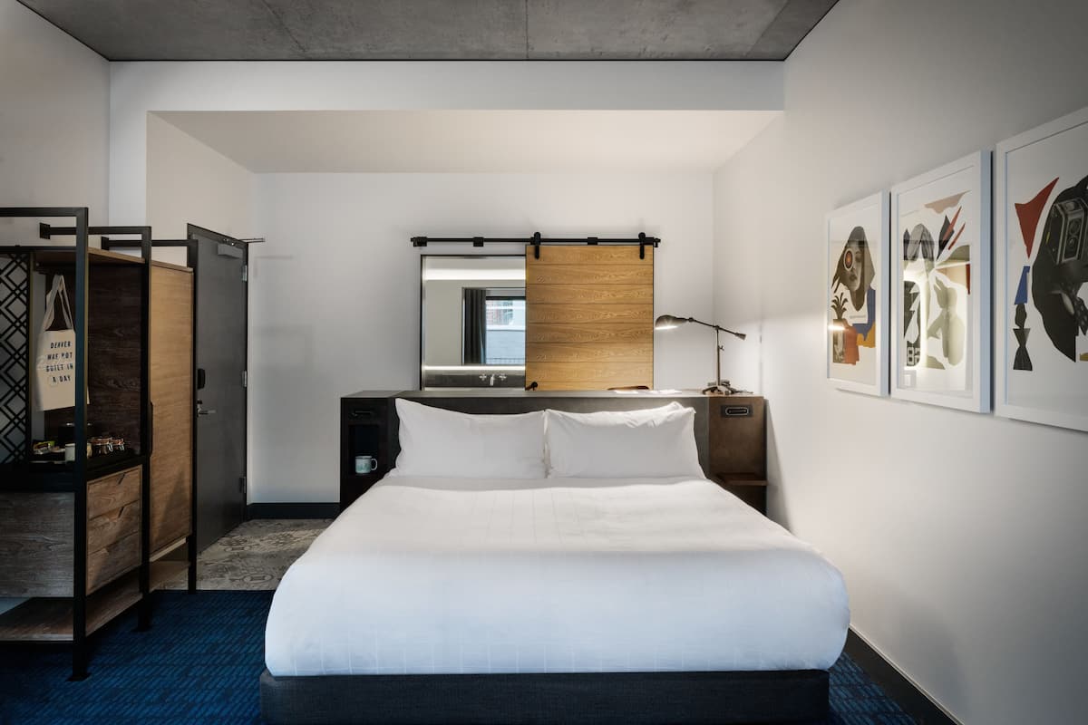 Quarto de hotel moderno com uma cama grande, um guarda-roupa de madeira, uma escrivaninha com abajur e quadros na parede.