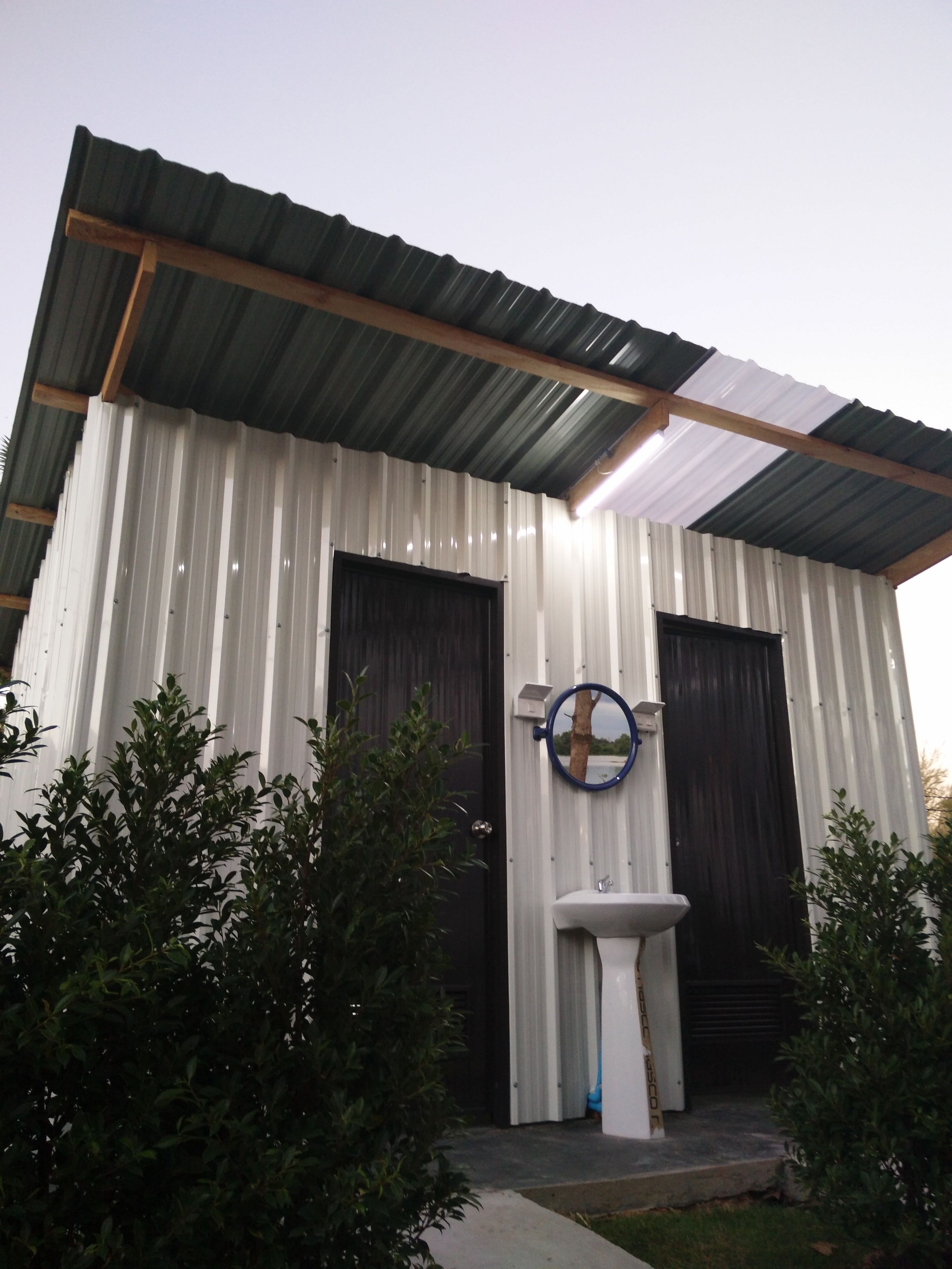 Um edifício com paredes de metal corrugado, duas portas escuras, uma pia branca e um pequeno jardim com arbustos.