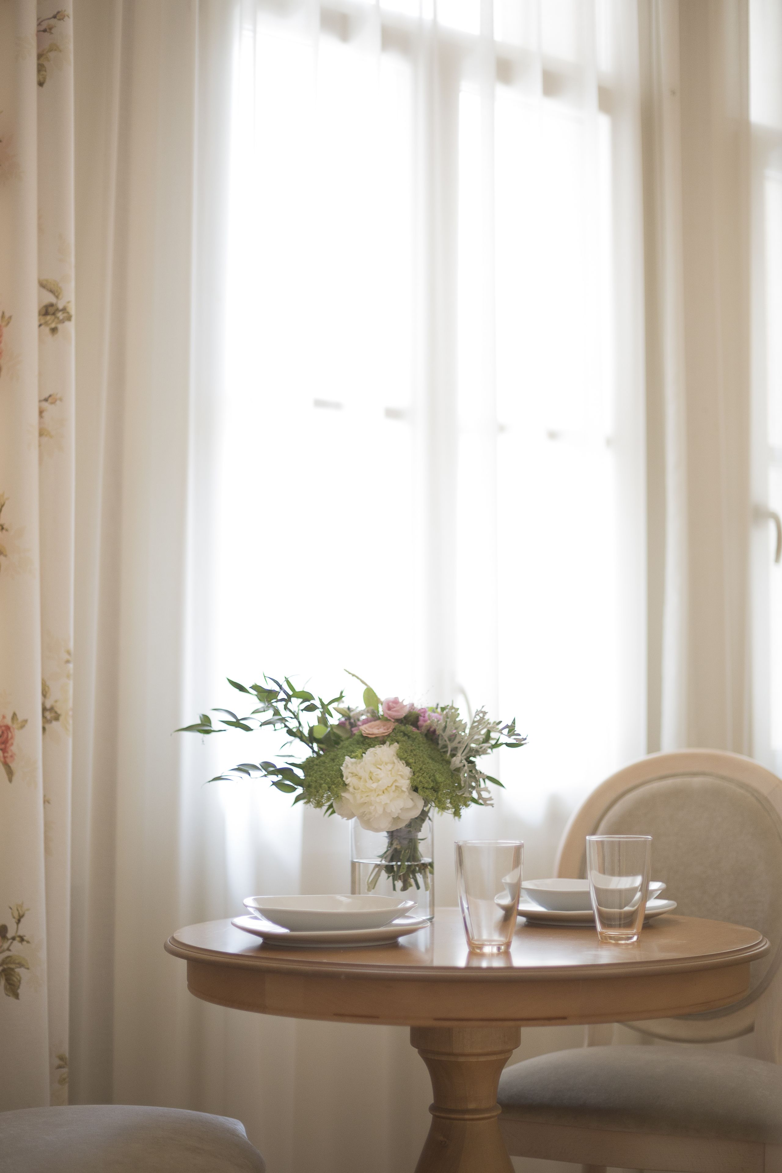 Mesa de jantar com um vaso de flores, duas taças e pratos, em frente a uma janela com cortinas translúcidas.