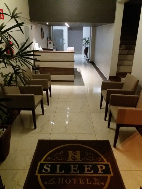 Uma área de recepção de hotel com balcão, algumas cadeiras e um grande vaso de plantas.