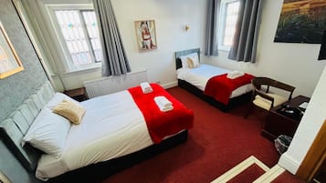 Quarto de hotel com duas camas, um tapete vermelho, um radiador, uma janela com cortinas, um quadro na parede e uma cadeira.