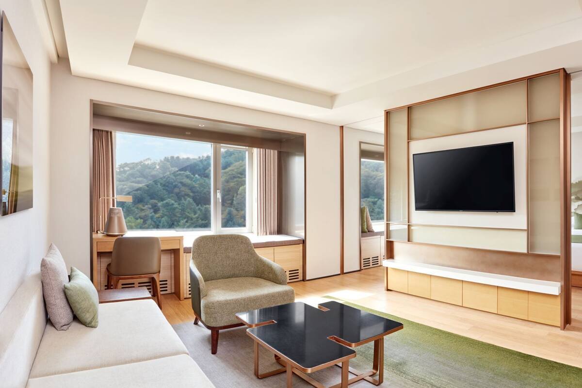 Quarto de hotel moderno com sofá, poltrona, televisão e escrivaninha com cadeira.