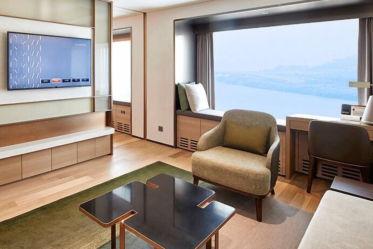 Quarto de hotel moderno com uma cama, uma televisão fixada na parede, um espaço de estar com uma poltrona e uma mesa, e uma grande janela com vista para o mar.