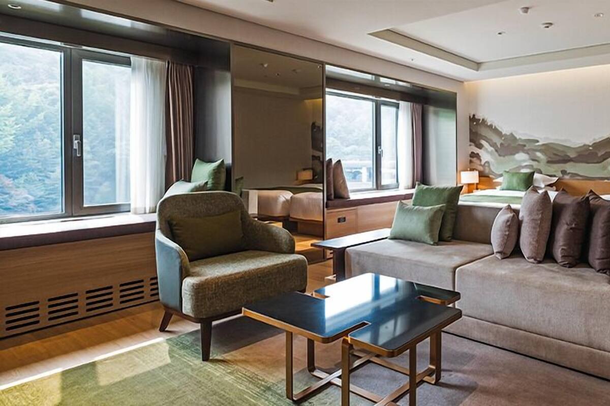 Quarto de hotel moderno com uma cama grande, um sofá, uma cadeira e uma pequena mesa. Há janelas com cortinas, um quadro na parede e vista para o exterior.