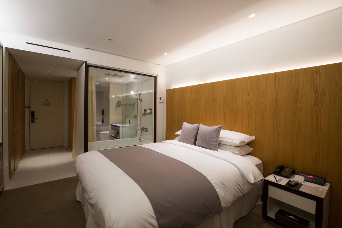 Quarto de hotel moderno com uma cama grande, cabeceira de madeira, mesa de cabeceira com telefone e relógio, e porta de vidro que dá para o banheiro.