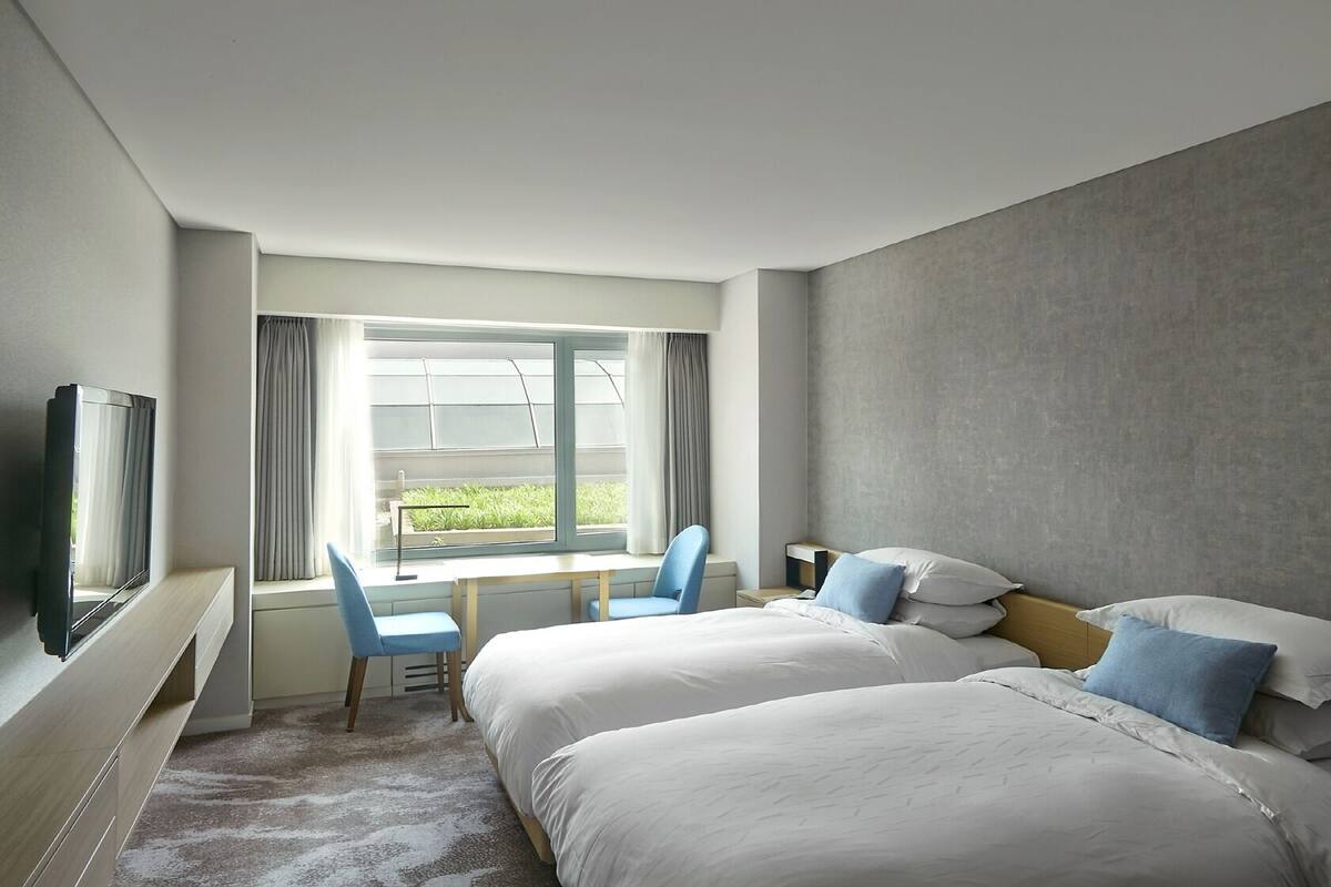 Quarto de hotel com duas camas, uma escrivaninha, cadeiras, uma televisão e uma janela com cortinas.