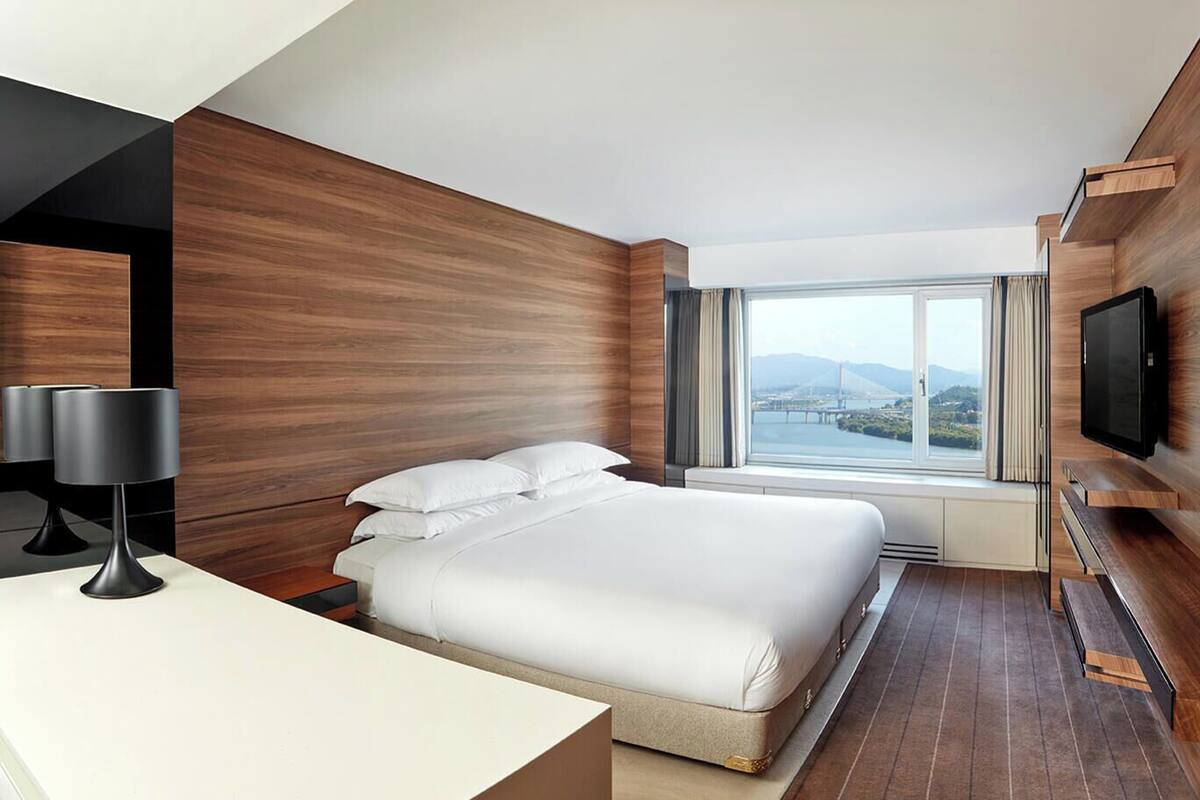 Quarto de hotel moderno com uma cama grande, uma escrivaninha, uma televisão e vista para uma ponte.