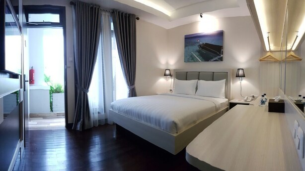 Quarto de hotel com uma cama grande, mesas de cabeceira, uma escrivaninha, uma cadeira, uma janela com cortinas, um quadro na parede e um extintor de incêndio no canto.