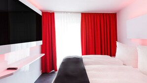 Quarto de hotel moderno com uma cama grande, televisão de tela plana na parede e cortinas vermelhas.