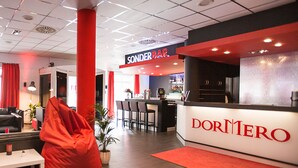 Um hall de hotel moderno com esquema de cores vermelho e branco, área de bar chamada 'Sonderbar' e balcão de recepção identificado como 'Dormiero'.
