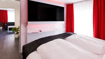 Quarto de hotel moderno com uma cama grande, uma TV de tela plana na parede branca, cortinas vermelhas e um vaso de planta.