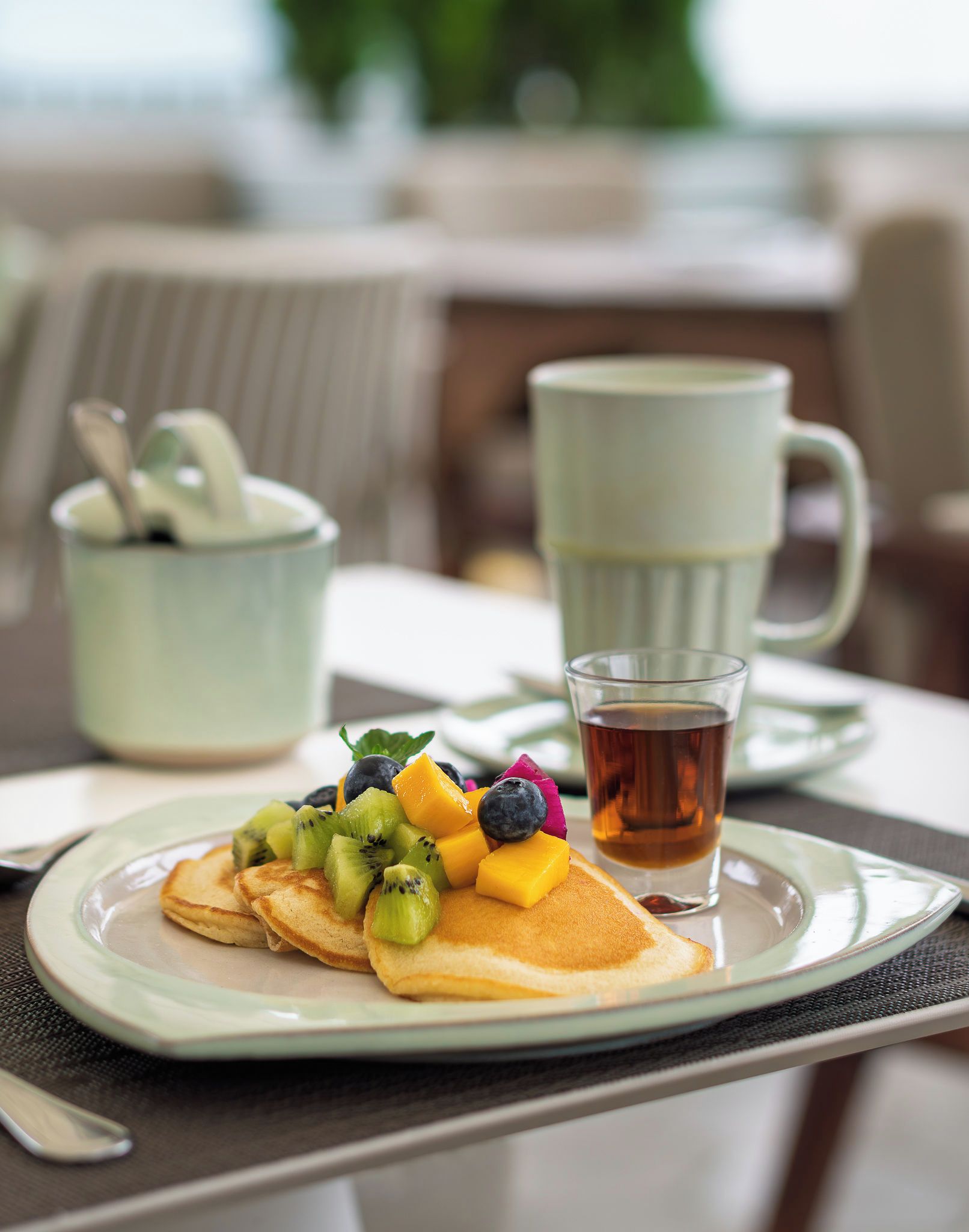 Disposição de café da manhã com panquecas, frutas e uma xícara de chá sobre uma mesa.