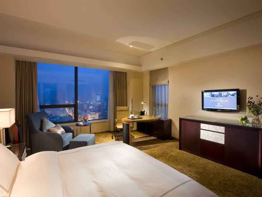 Quarto de hotel com uma cama grande, uma escrivaninha com cadeira, televisão e vista para a cidade ao entardecer.