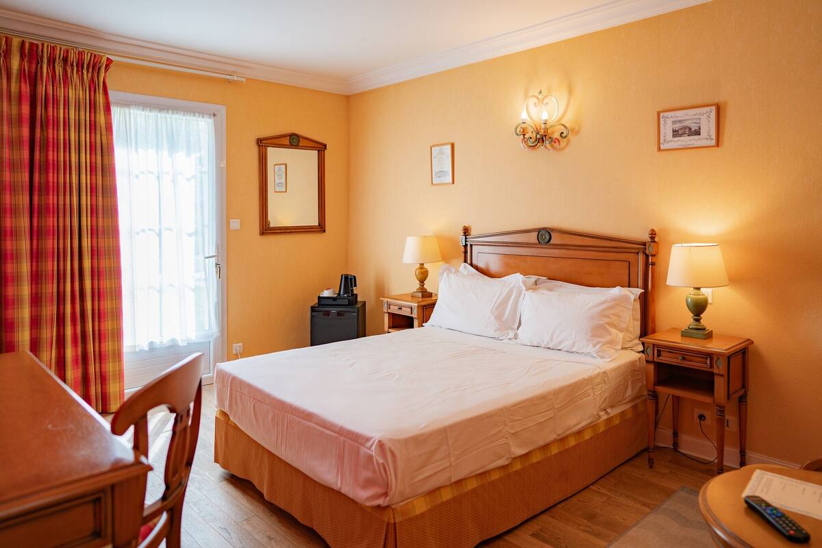 Quarto de hotel com uma cama grande, duas mesas de cabeceira com luminárias, uma escrivaninha com cadeira e uma janela com cortinas.