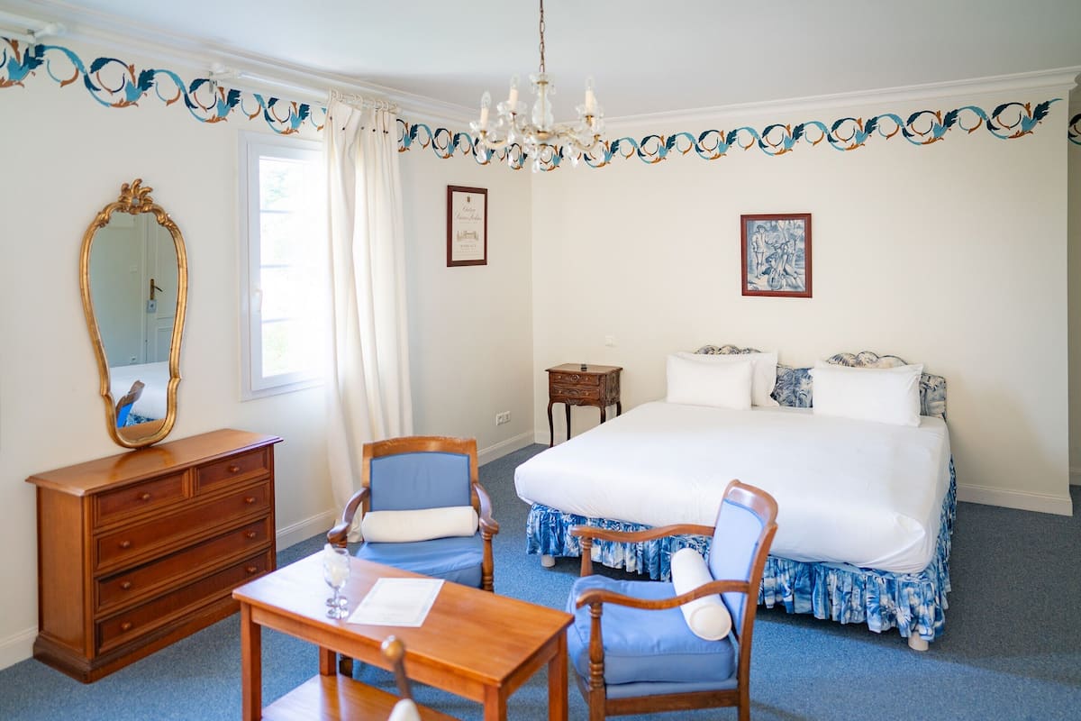 Quarto com cama grande, duas cadeiras, penteadeira de madeira, mesa, espelho e lustre.