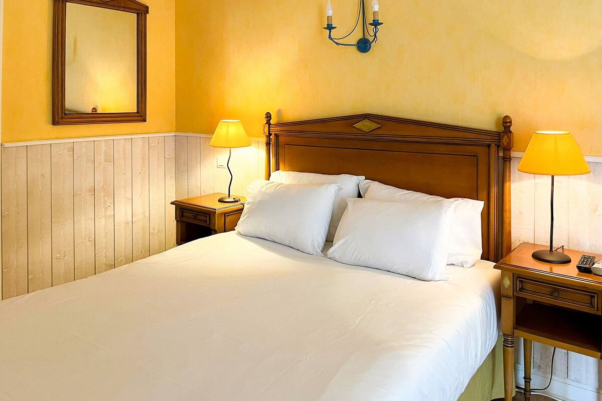 Cama bem arrumada com roupa de cama branca, duas mesas de cabeceira com luminárias, cabeceira de madeira e um espelho na parede.