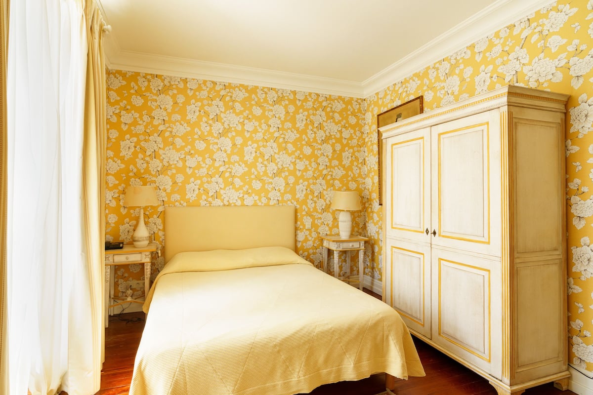 Um quarto com uma cama, duas mesas de cabeceira com abajures, um guarda-roupa e papel de parede floral.