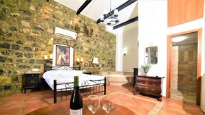 Quarto com parede de pedra, uma cama, cômoda, espelho e uma garrafa de vinho sobre uma mesa.