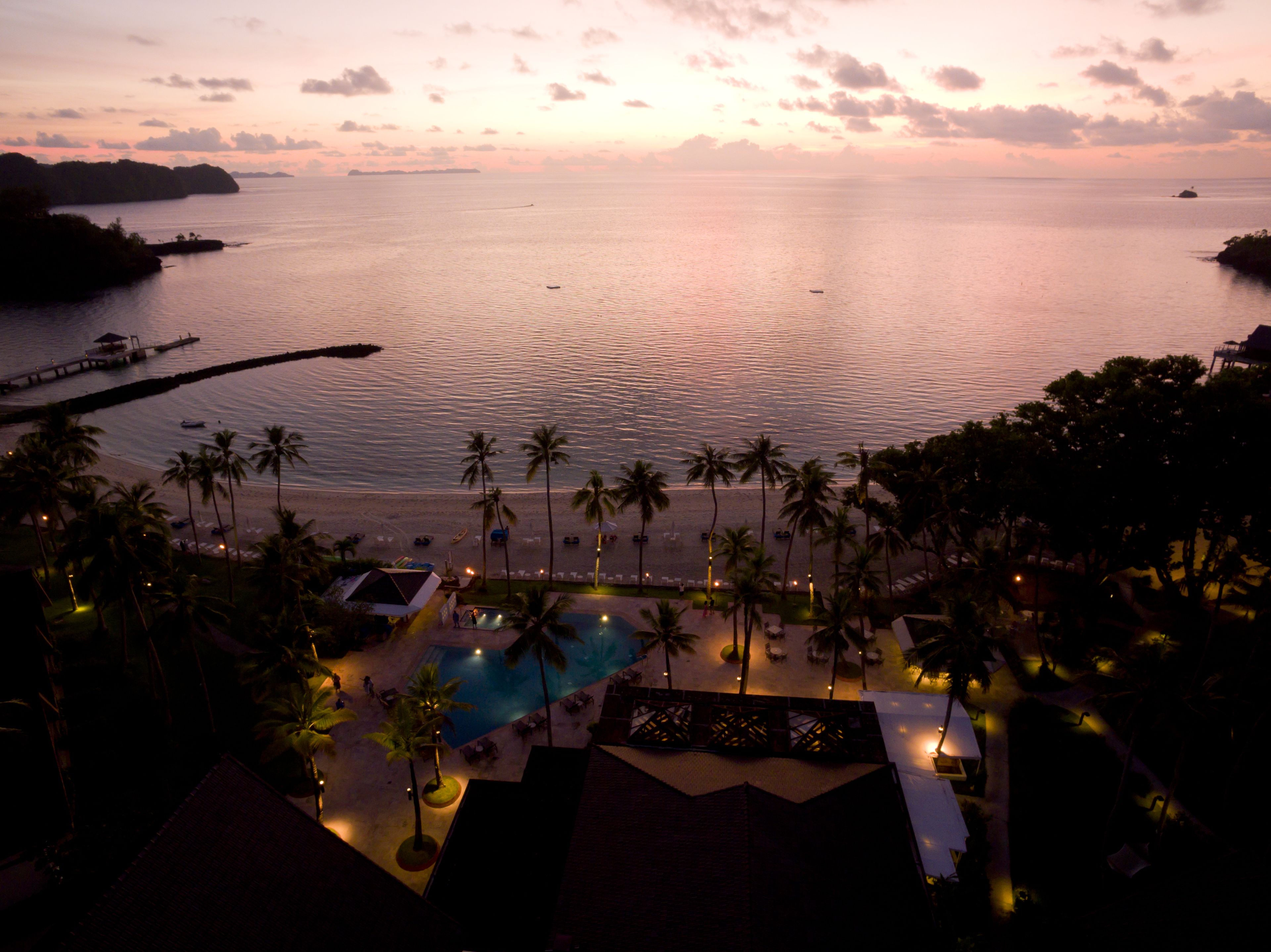 Resort com piscina, palmeiras e vista para o oceano ao pôr do sol.