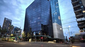 Um edifício moderno de arranha-céu com fachada de vidro, rodeado por outros edifícios altos e uma linha de elétrico.