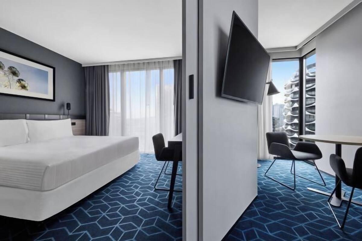 Quarto de hotel com uma cama, uma escrivaninha, duas cadeiras, uma televisão e uma janela grande.