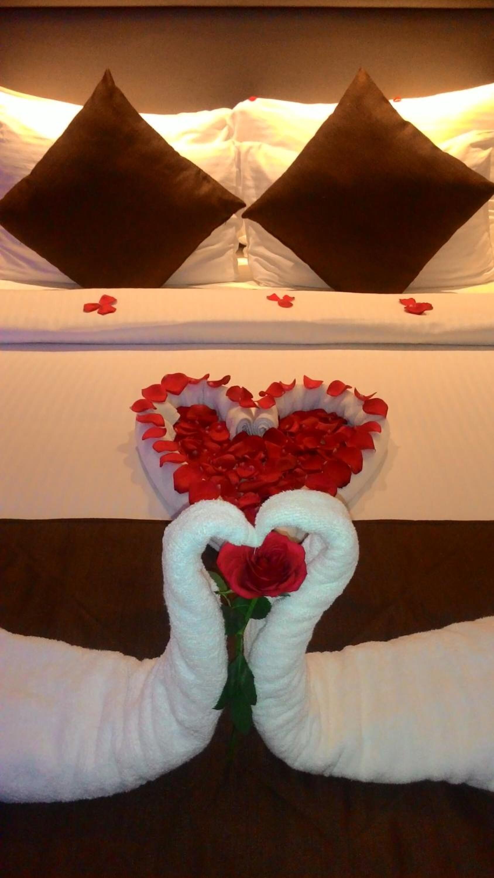 Uma cama com um arranjo de pétalas de rosas vermelhas em formato de coração e uma rosa, além de duas toalhas moldadas como cisnes.