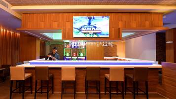 Uma área de bar com uma televisão exibindo 'GAME PLAN' e 'TESTS'. Há banquetas de bar e um balcão.