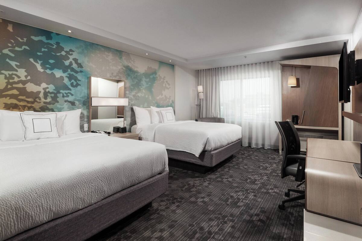 Quarto de hotel com duas camas, uma escrivaninha, uma cadeira, televisão e uma janela grande com cortinas.