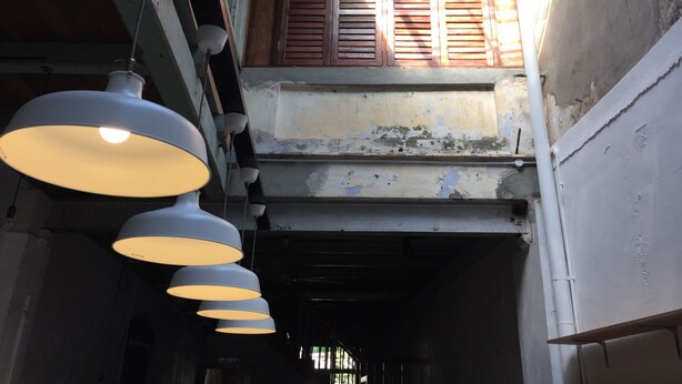 Uma viela estreita com várias luminárias pendentes, uma janela com persianas de madeira e uma placa visível para 'KELIART'.