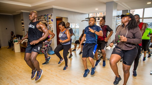 Um grupo de pessoas se exercitando em uma sala com piso de madeira e equipamentos de ginástica.