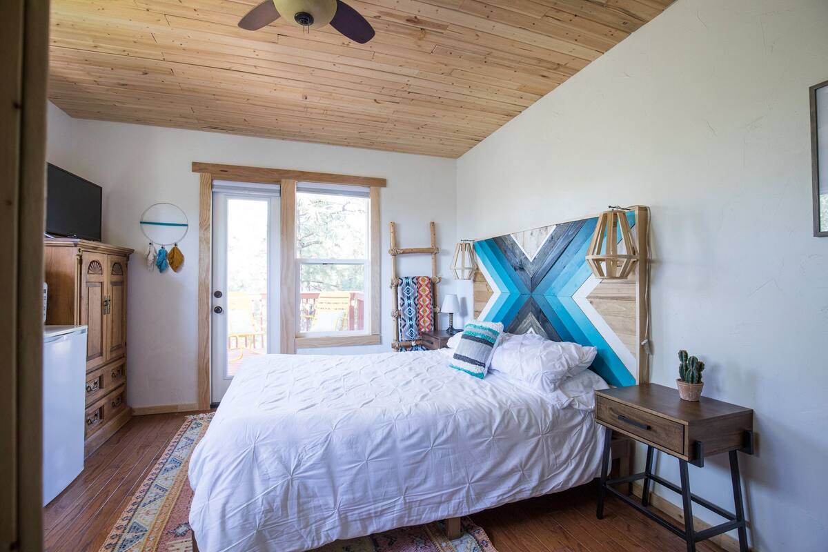 Quarto com cama, teto de madeira, escrivaninha, cadeira, televisão e janela.