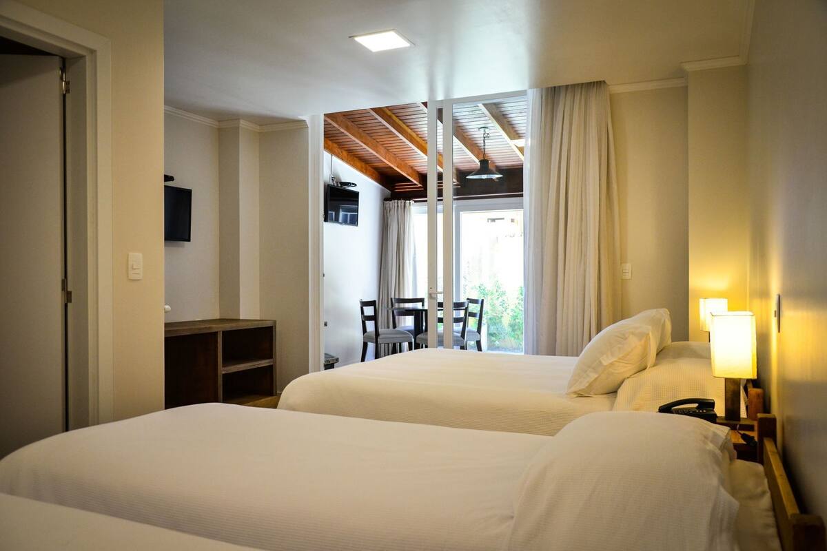 Quarto de hotel com duas camas, uma escrivaninha, uma cadeira e uma janela com cortinas.